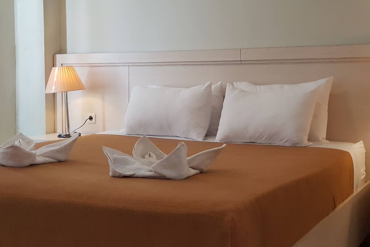 Cama bem arrumada com almofadas brancas e manta marrom, abajur e um travesseiro decorativo em formato de cisne na cama.