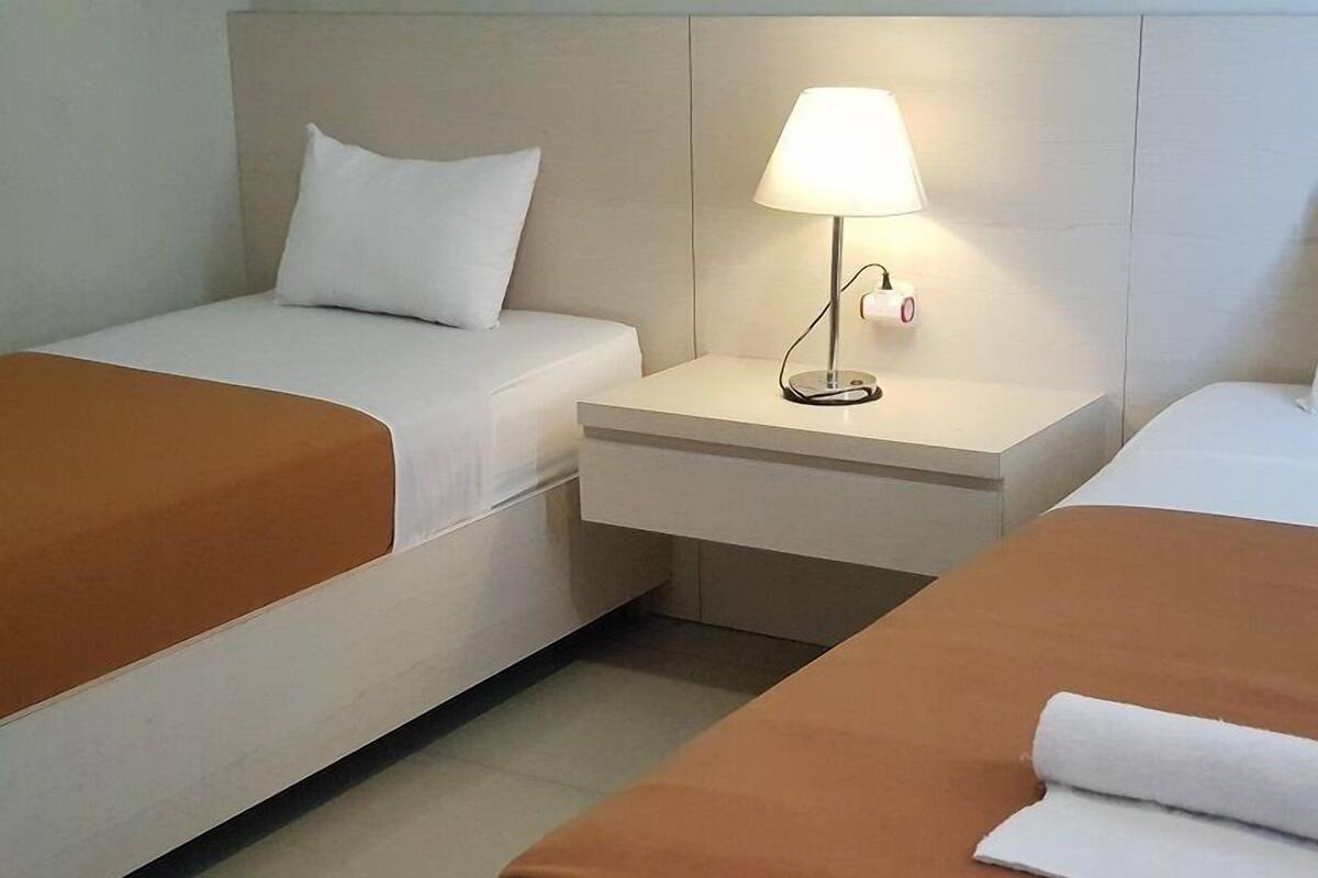 Quarto de hotel com duas camas, uma mesa de cabeceira com abajur e um travesseiro branco.
