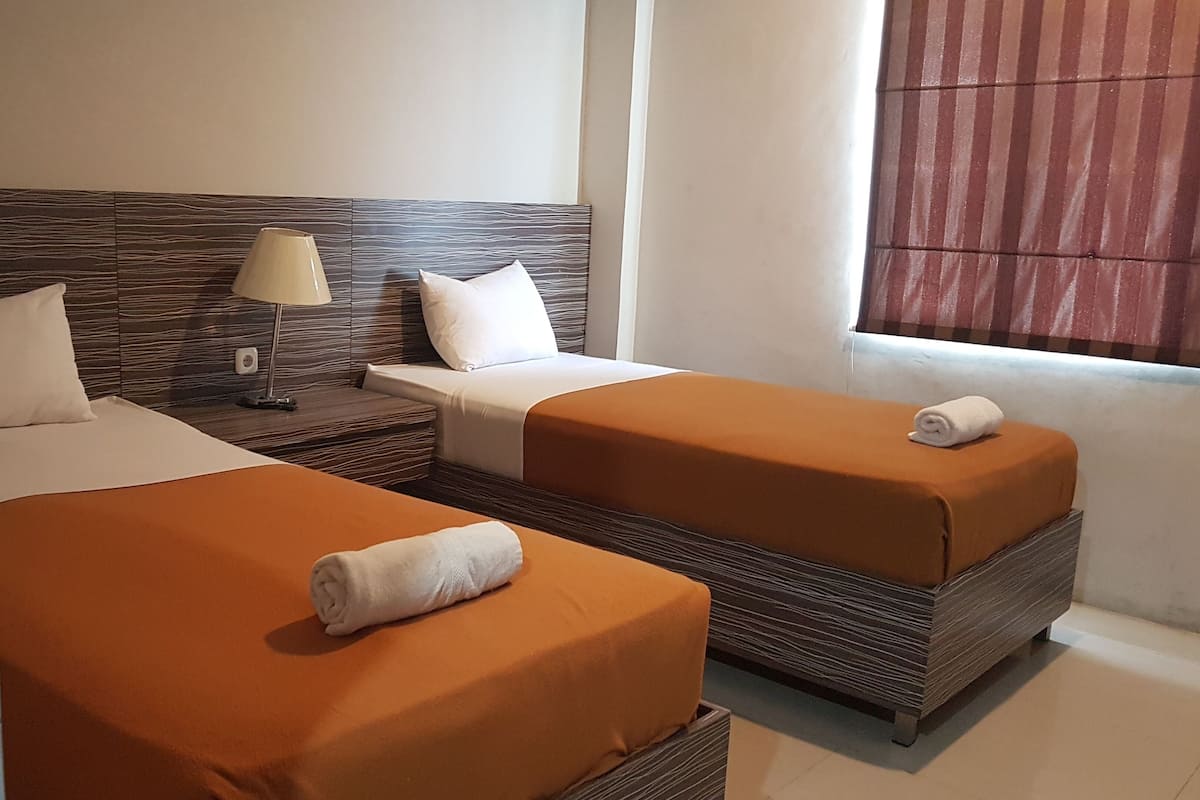 Quarto de hotel com duas camas de solteiro, mesa de cabeceira com abajur e janela com cortinas.