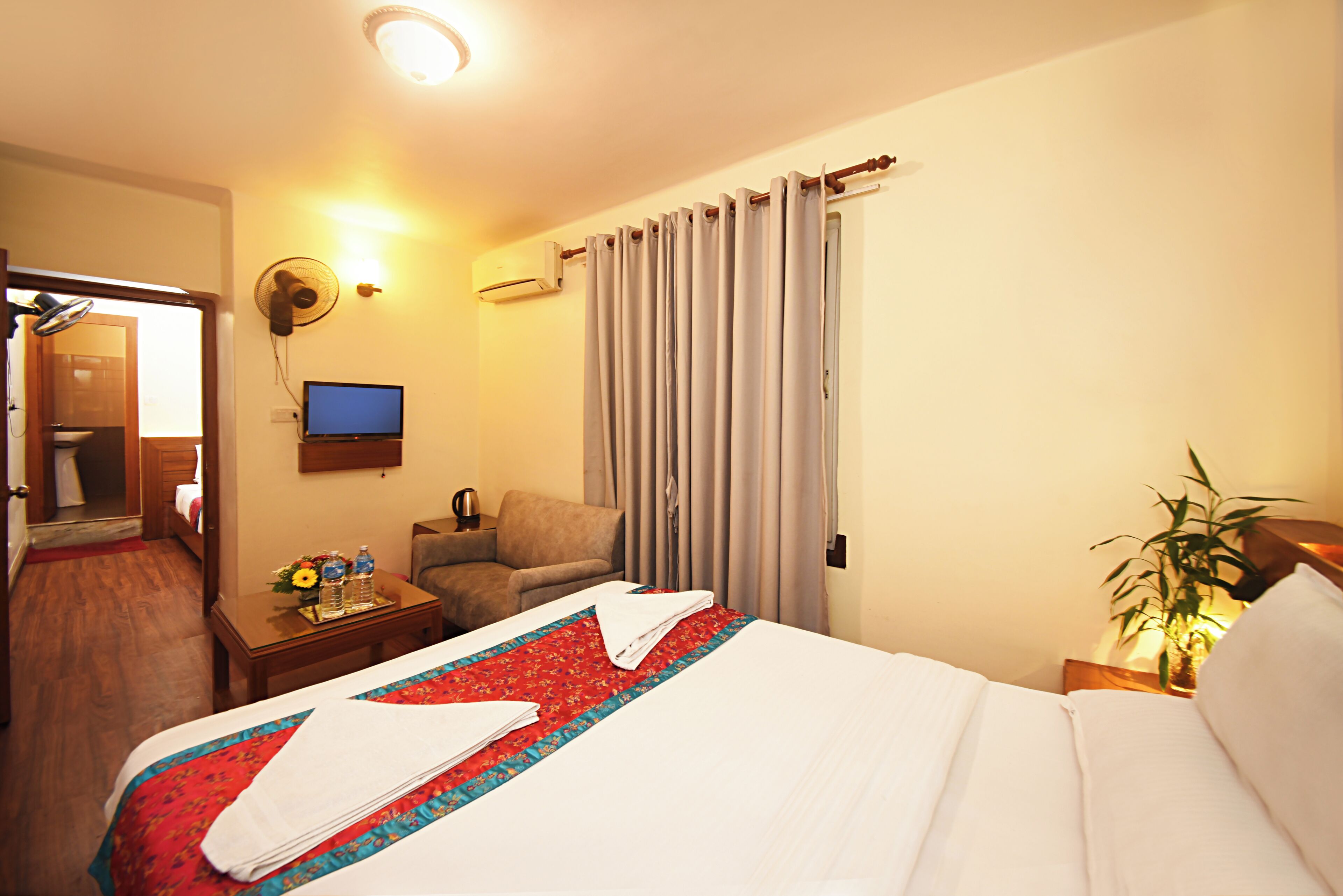 Quarto de hotel com cama, escrivaninha com luminária, cadeira, televisão e janela com cortinas.