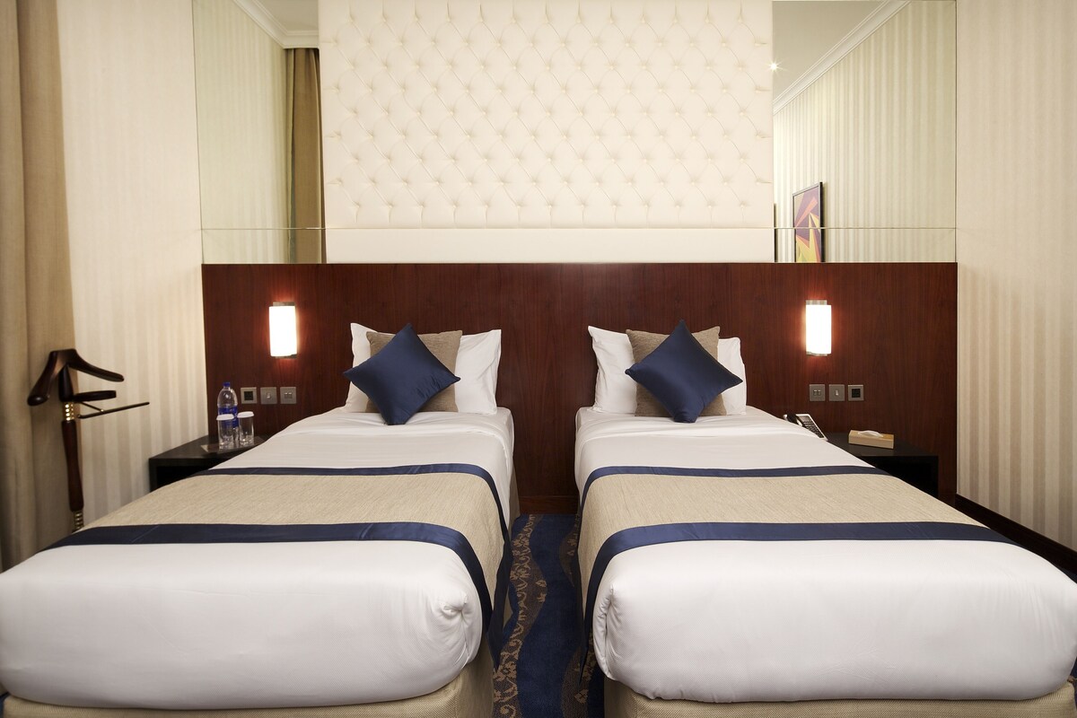 Quarto de hotel com duas camas, cabeceira de madeira, luminárias de cabeceira, espelho e mesa de cabeceira com telefone e garrafa.