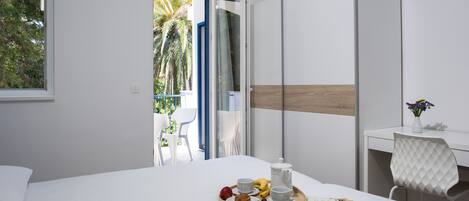 Quarto de hotel com uma cama, uma mesa de jantar com itens de café da manhã, uma cadeira branca e vista para a varanda.