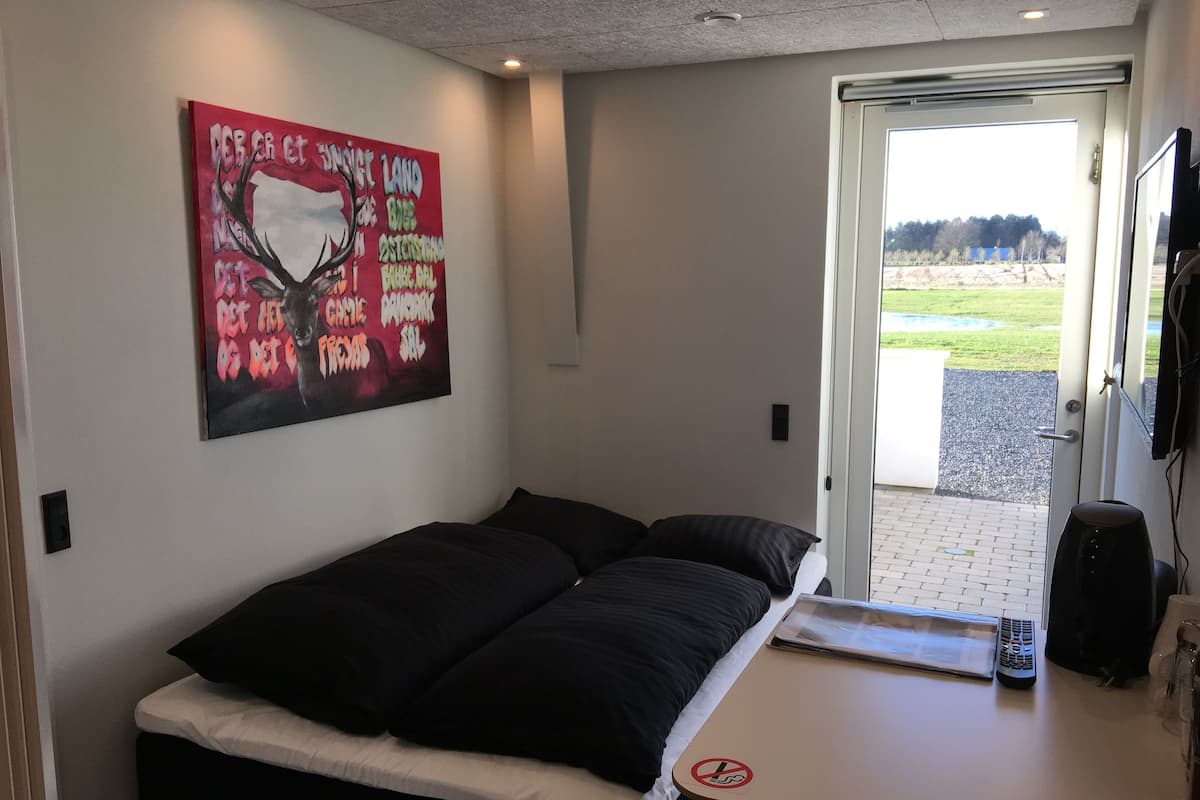 Quarto com cama, uma mesinha, televisão e porta que dá para o exterior.