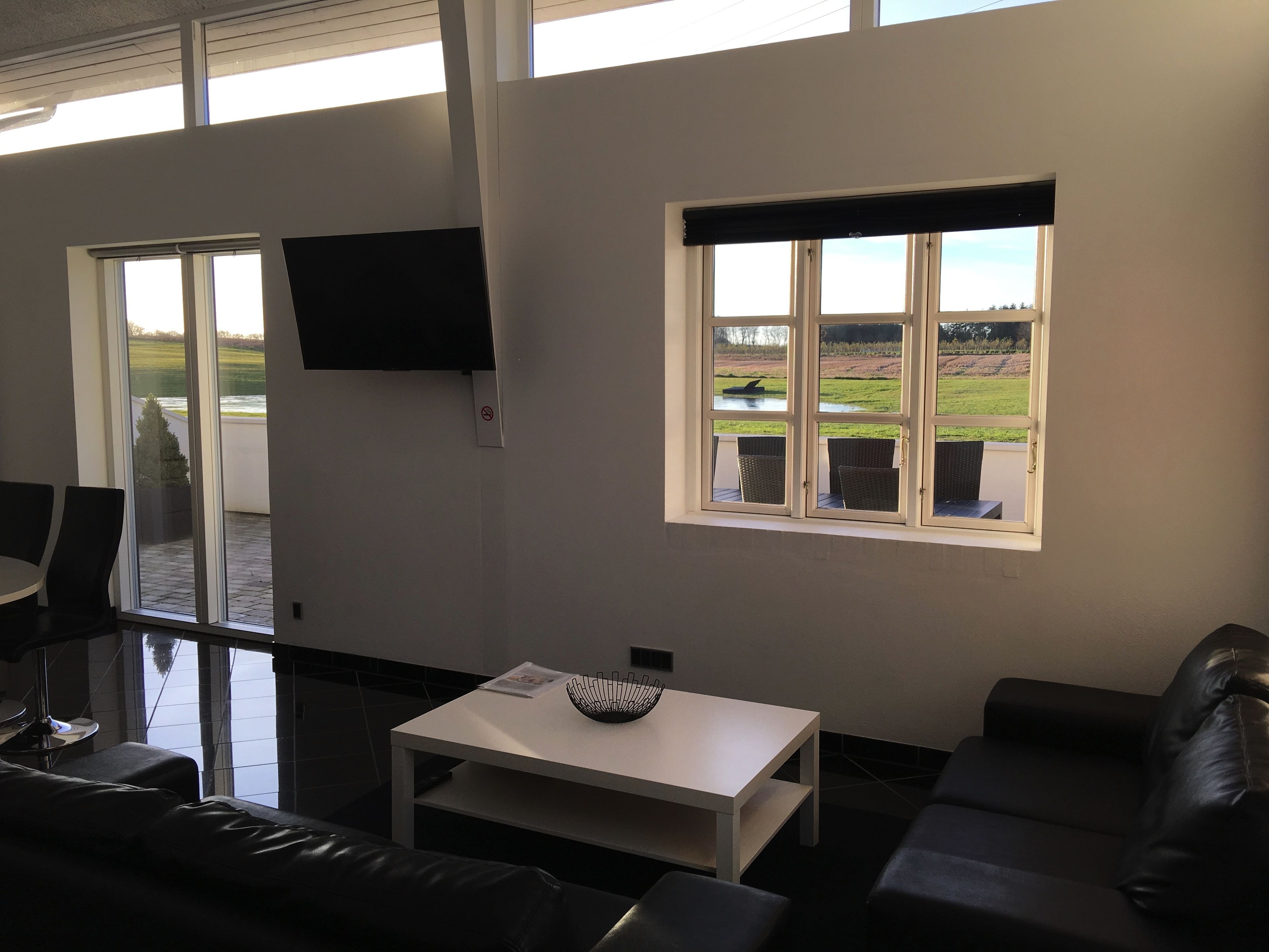 Uma sala de estar moderna com uma mesa de centro de vidro, um sofá branco e uma janela com vista para um campo gramado.