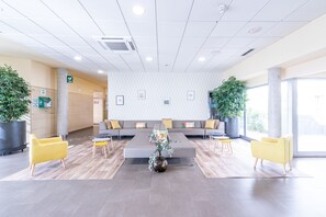 Uma área de espera moderna com um sofá grande, cadeiras amarelas, uma mesa de centro e plantas em vasos.