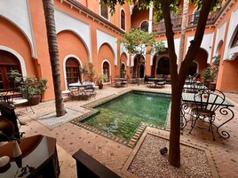 Um pátio externo com piscina central, rodeado por mesas e cadeiras, e palmeiras.