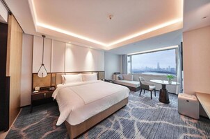 Quarto de hotel moderno com uma cama grande, área de estar, uma mesinha e vista para o horizonte da cidade.