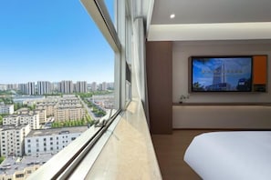 Quarto de hotel com uma janela grande, uma cama, uma TV e vista para a paisagem urbana.