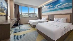 Quarto de hotel com duas camas, uma escrivaninha, uma cadeira, uma janela grande com vista para a cidade e uma pintura de uma cena de praia.
