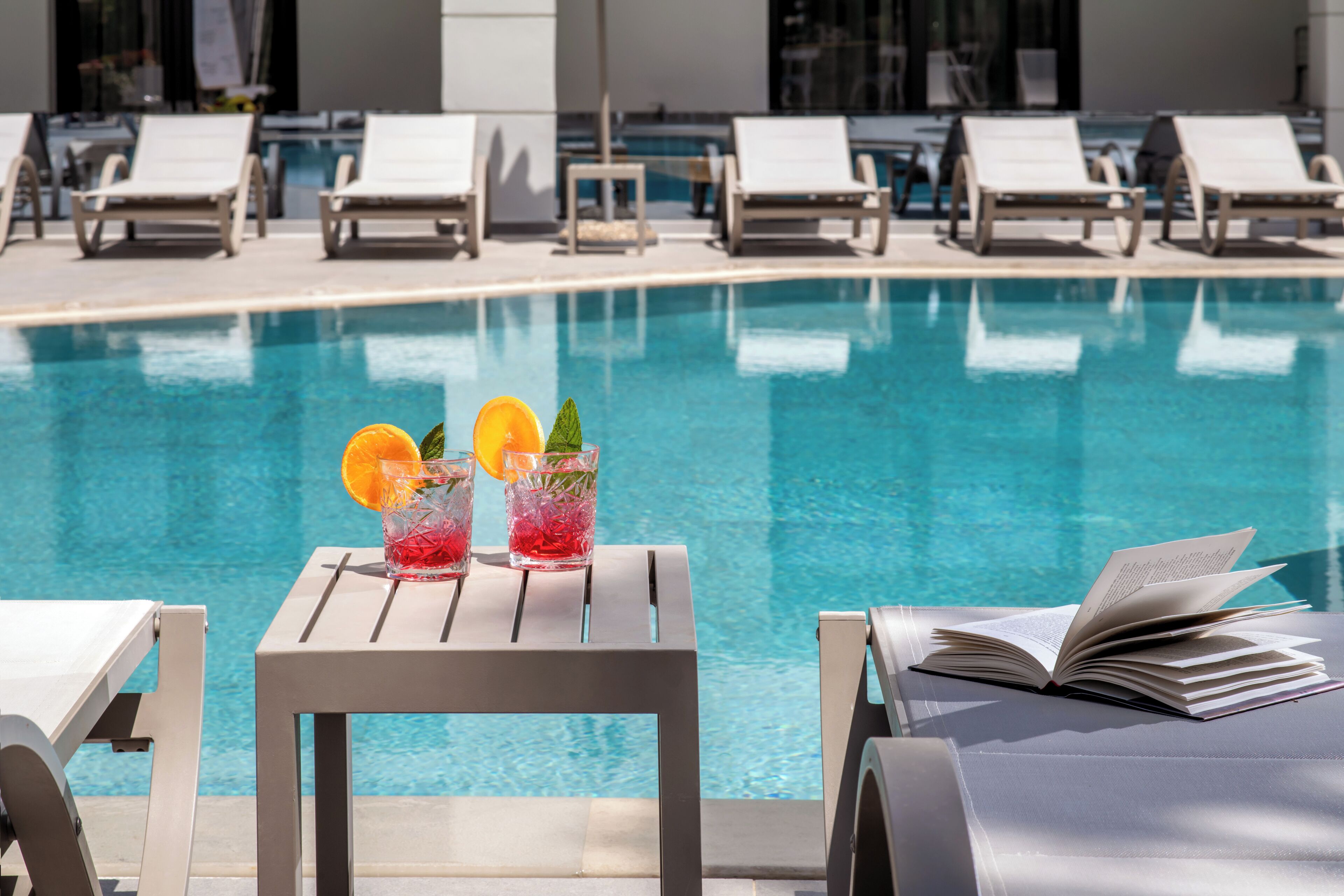 Uma área à beira da piscina com espreguiçadeiras, uma mesa com bebidas e um livro aberto.