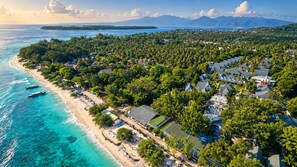 Vista aérea de um resort costeiro com propriedades na orla, águas turquesas cristalinas e montanhas ao fundo.