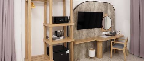 Quarto de hotel compacto com escrivaninha de madeira, televisão, espelho, armário pequeno e sapateira.