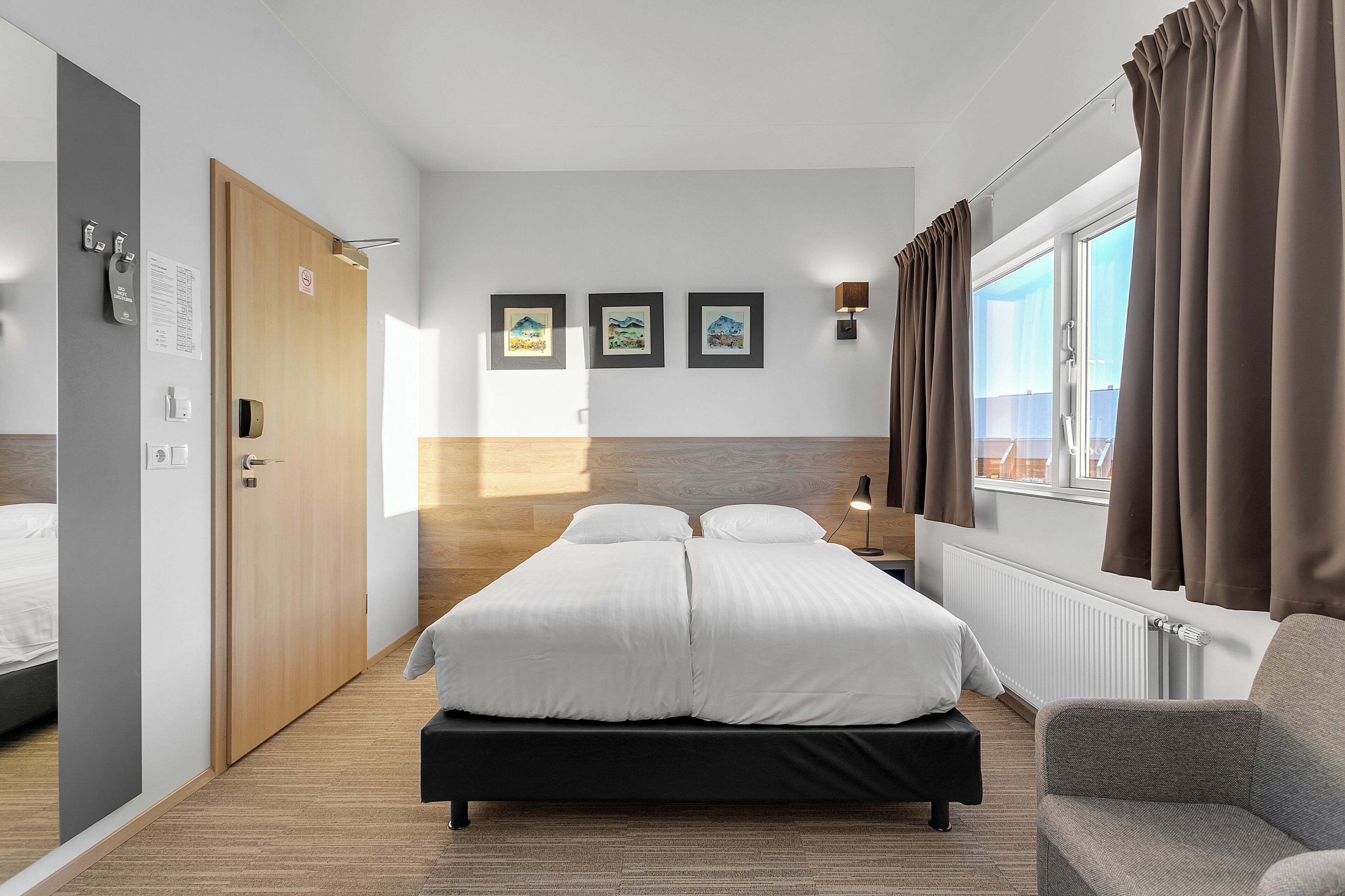 Quarto de hotel com cama, poltrona, janela com cortinas e três quadros na parede.
