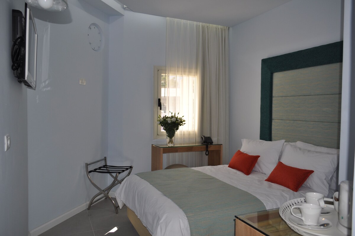 Quarto de hotel com cama, escrivaninha com telefone, cadeira, janela com cortinas e televisão fixada na parede.