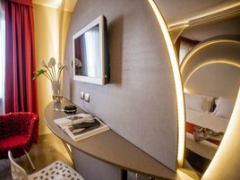 Quarto de hotel moderno com escrivaninha curva, cama com roupa de cama branca, uma cadeira vermelha e televisão fixada na parede.