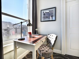 Quarto com escrivaninha, cadeira, janela com vista para um edifício, quadro na parede e abajur.