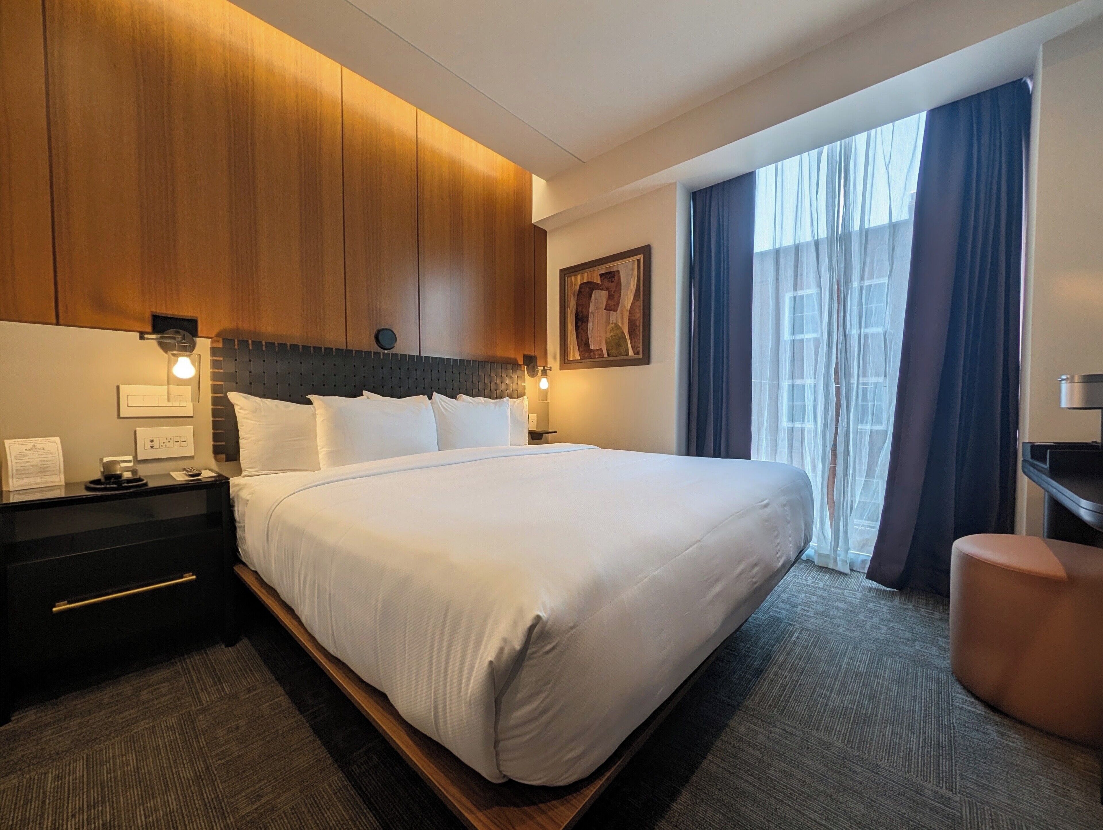 Quarto de hotel com uma cama grande, cabeceira de madeira, abajures, uma escrivaninha e uma janela ampla com cortinas.