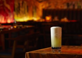 Um copo alto de uma bebida cremosa sobre uma mesa de madeira em um bar com pouca luz, móveis de madeira e fundo desfocado.