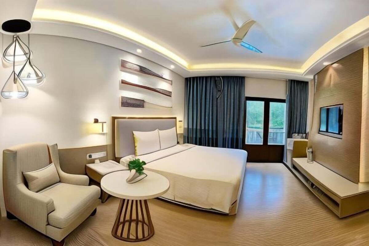 Quarto de hotel com uma cama grande, um sofá, uma mesinha redonda, uma televisão e um ventilador de teto.