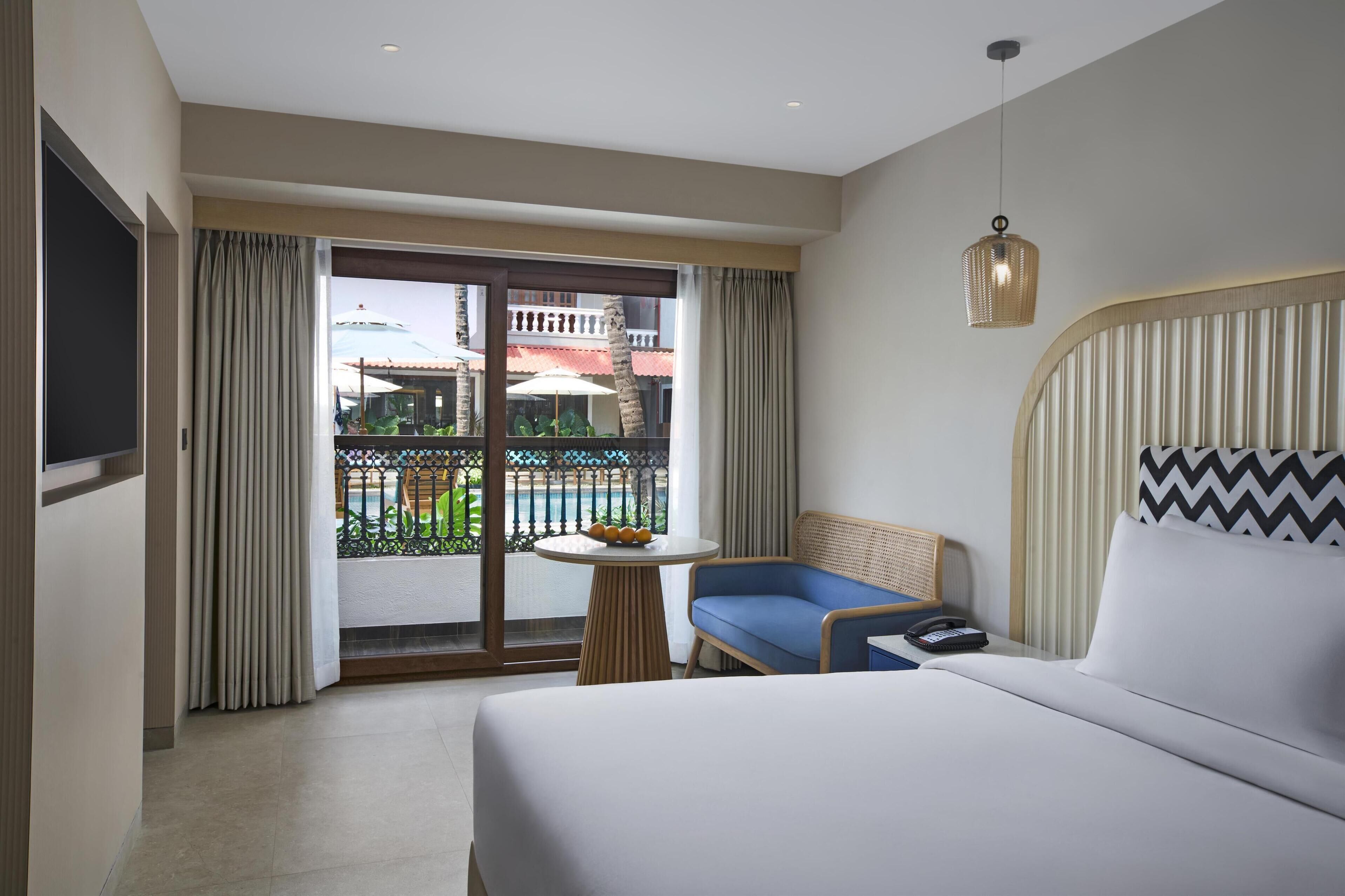 Quarto de hotel com uma cama, uma mesinha com frutas, uma cadeira azul, uma varanda com vista para prédios e uma piscina, e um lustre.