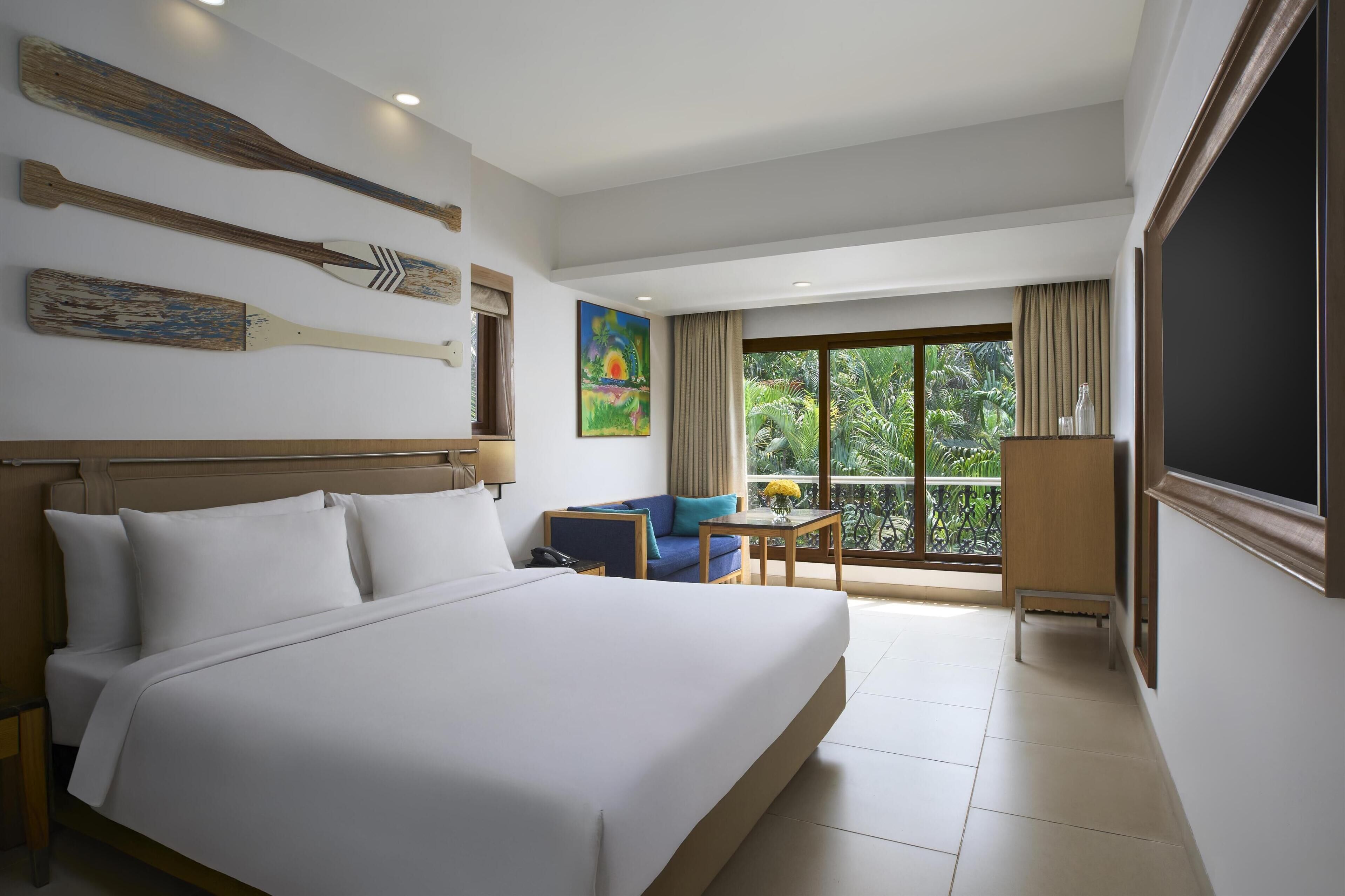 Quarto de hotel com uma cama grande, uma escrivaninha, uma cadeira, varanda com vista para o verde e um quadro na parede.