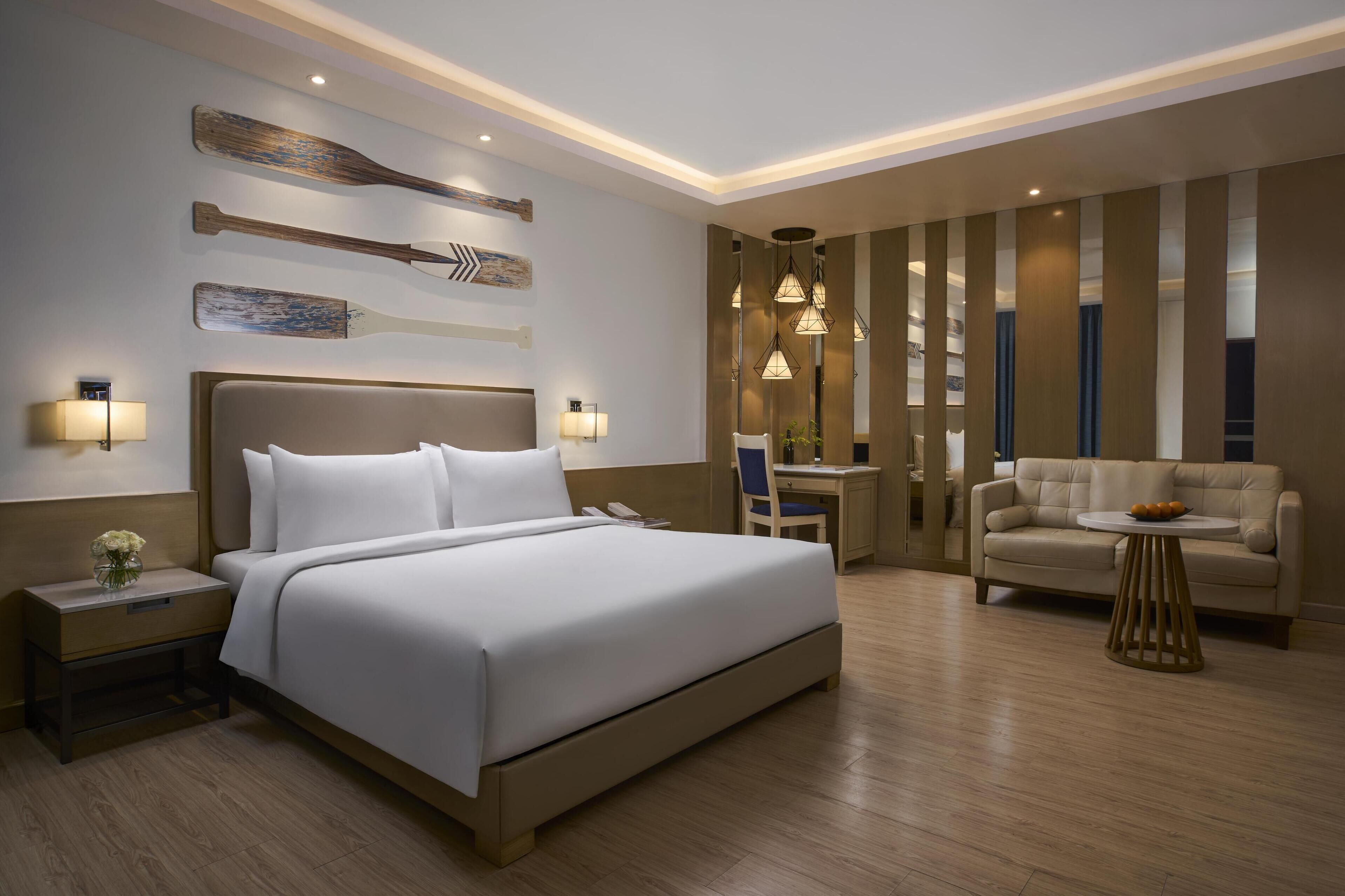 Quarto de hotel moderno com uma cama grande, mesas de cabeceira, escrivaninha, cadeira e sofá.