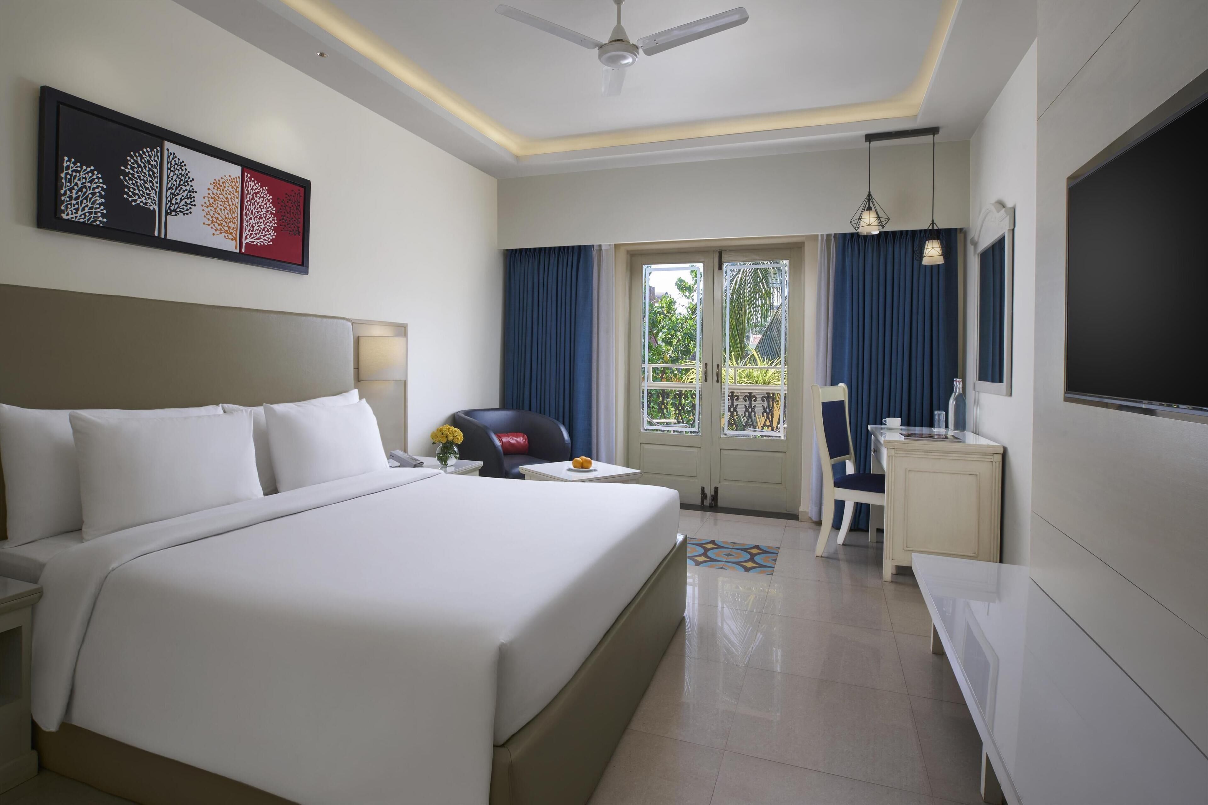 Quarto de hotel com uma cama grande, escrivaninha, cadeira, televisão, ventoinha no teto e varanda com cortinas.