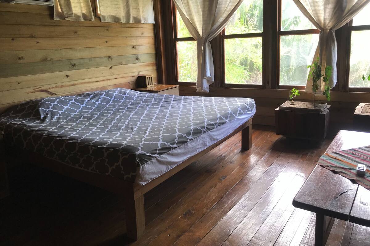 Quarto de cabana de madeira com cama, uma mesinha e uma janela com cortinas.