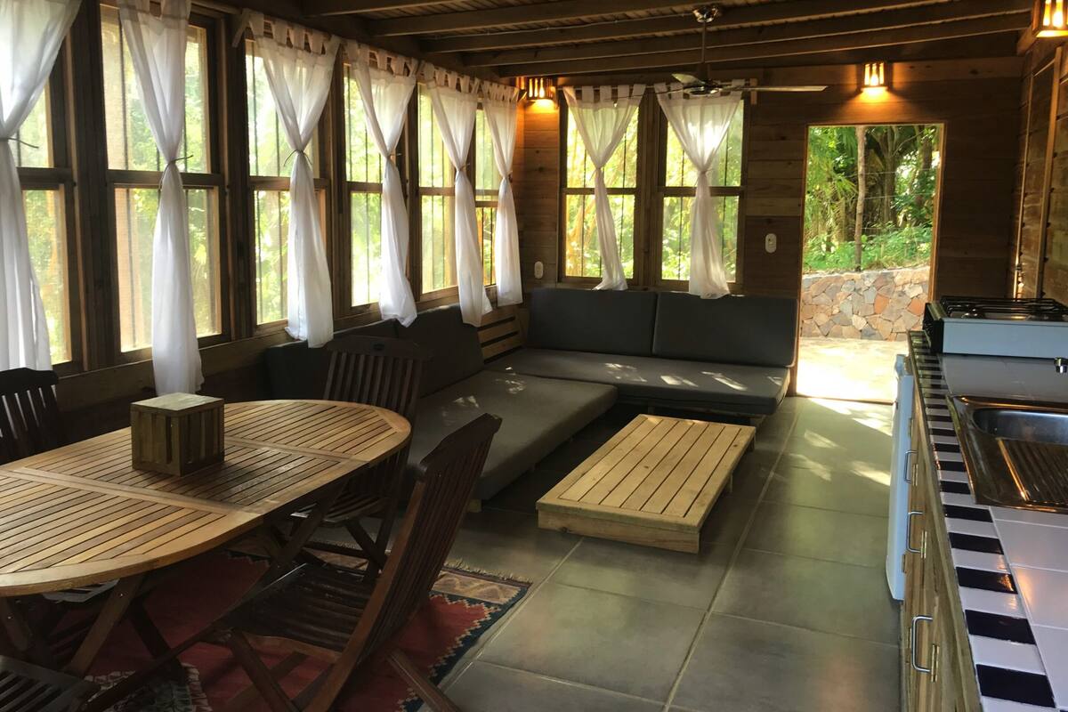 Interior acolhedor de uma cabana, com teto de madeira, sala de jantar com mesa redonda e cadeiras, área de estar com sofá e almofadas, e cozinha com fogão.