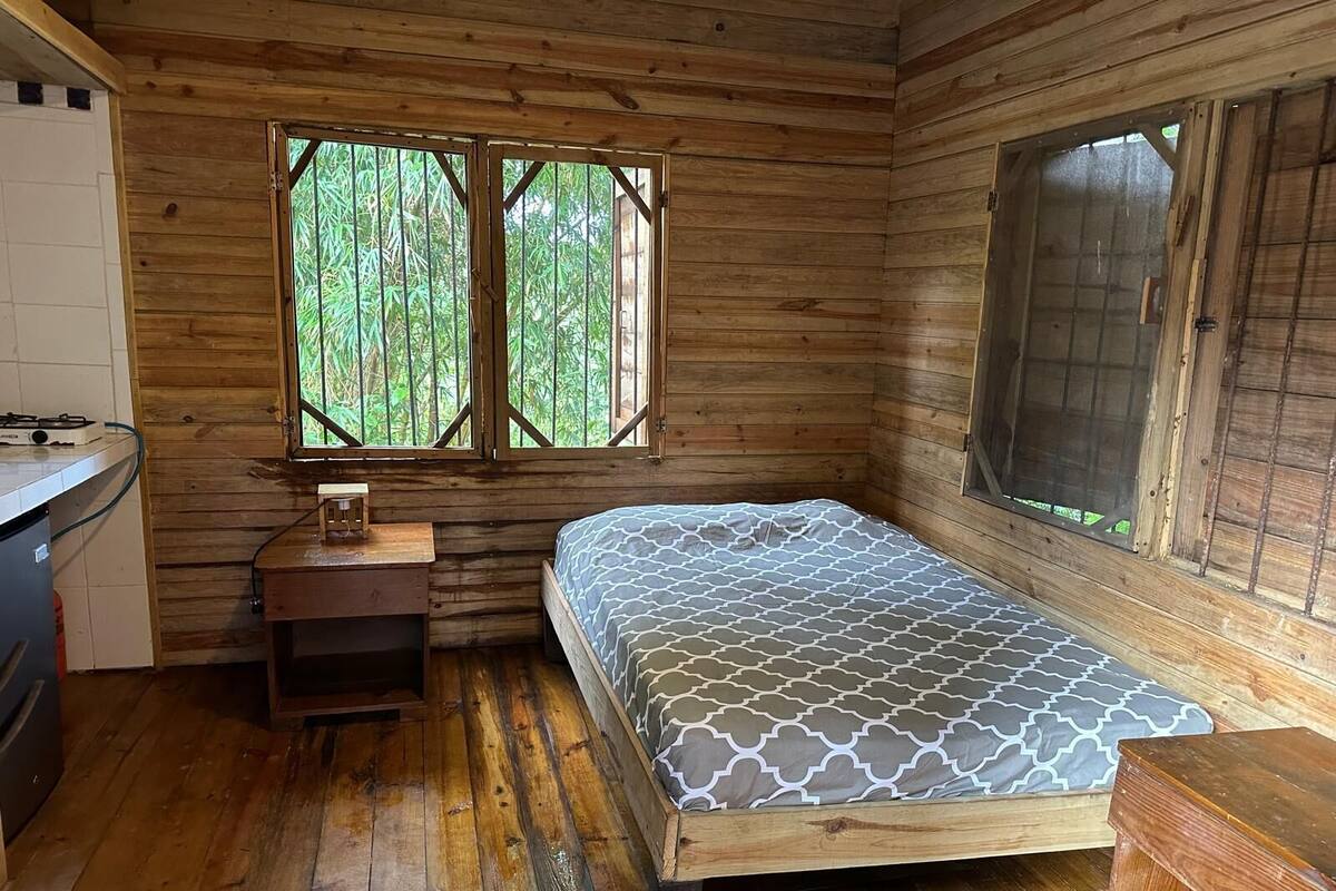 Quarto de cabana de madeira com cama, mesa pequena, janela e vista para o verde lá fora.