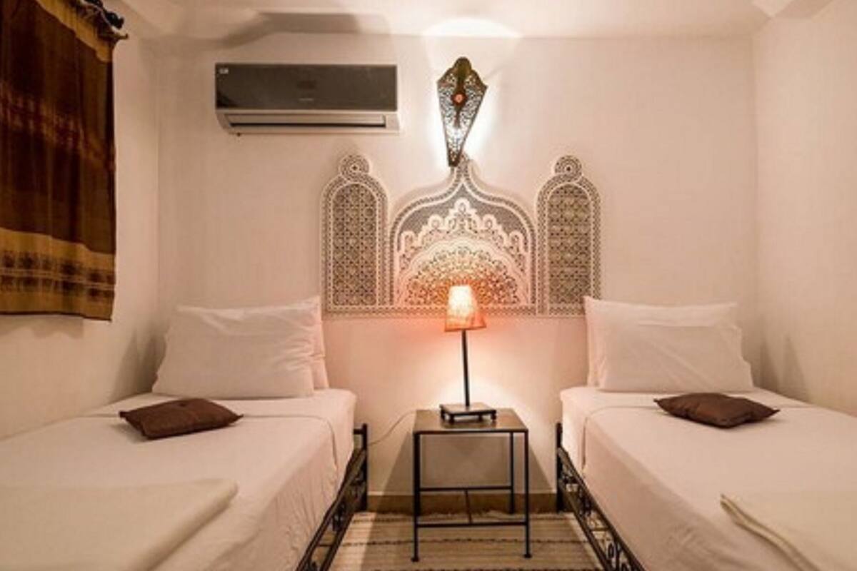 Quarto de hotel com duas camas, uma mesa de cabeceira com abajur e painel decorativo na cabeceira da cama.