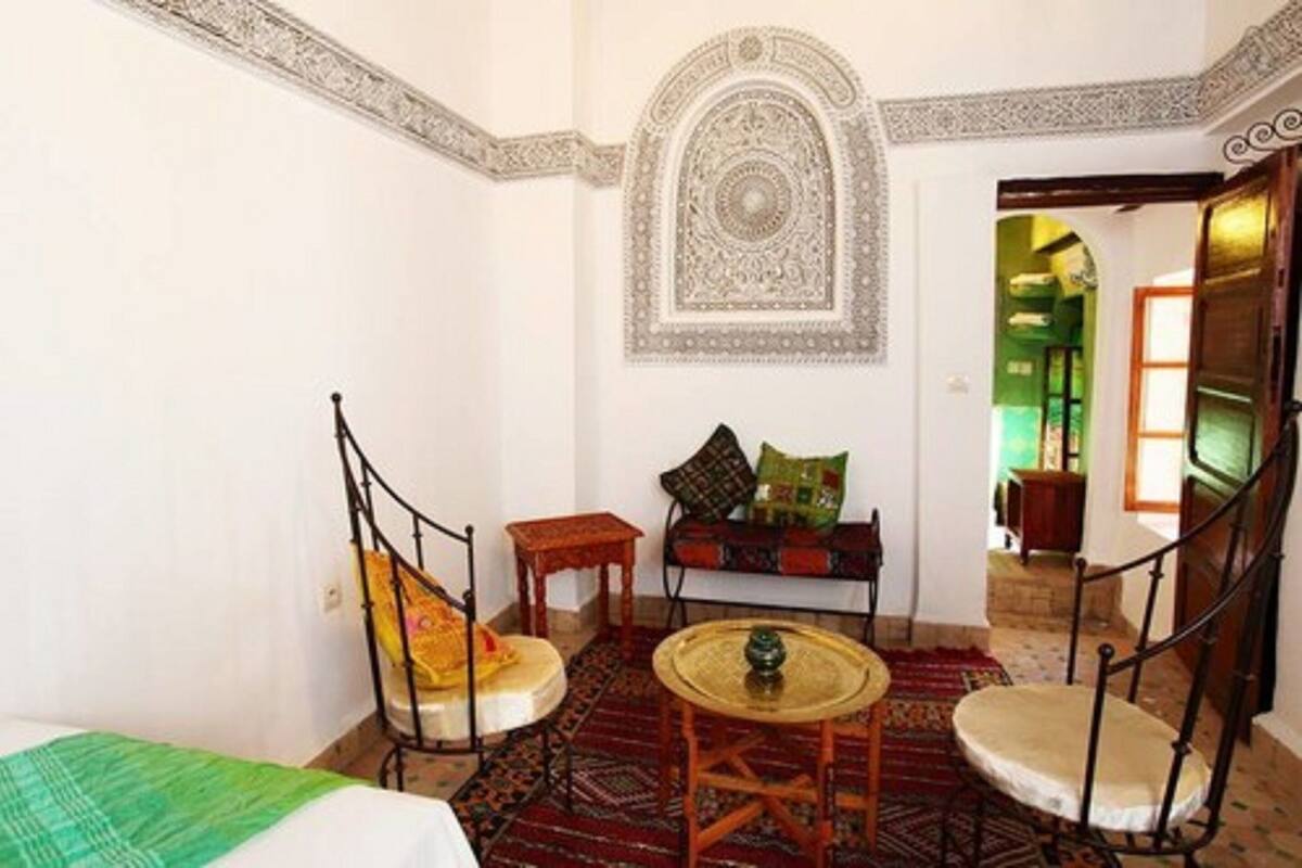 Quarto com cama, duas cadeiras, uma mesinha, um objeto decorativo na parede e um tapete estampado.