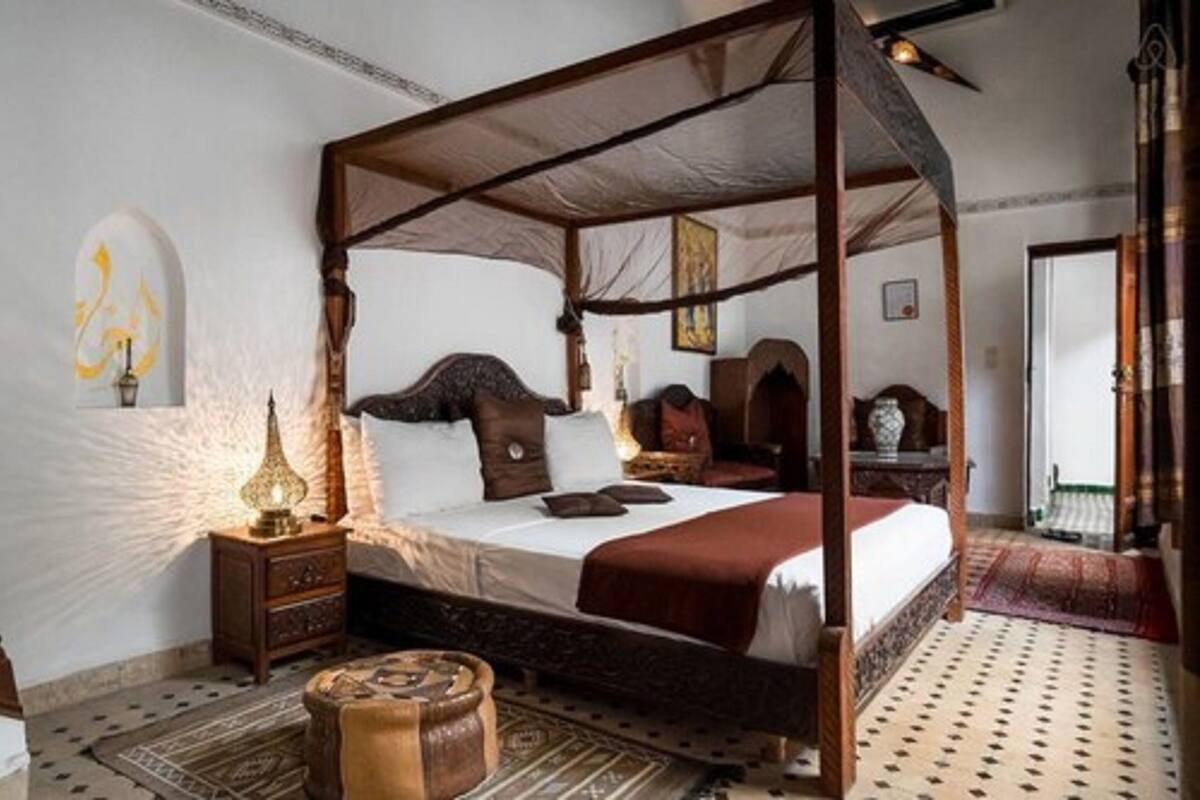 Uma cama com dossel e cortina, uma mesinha de cabeceira com abajur e um tapete estampado.