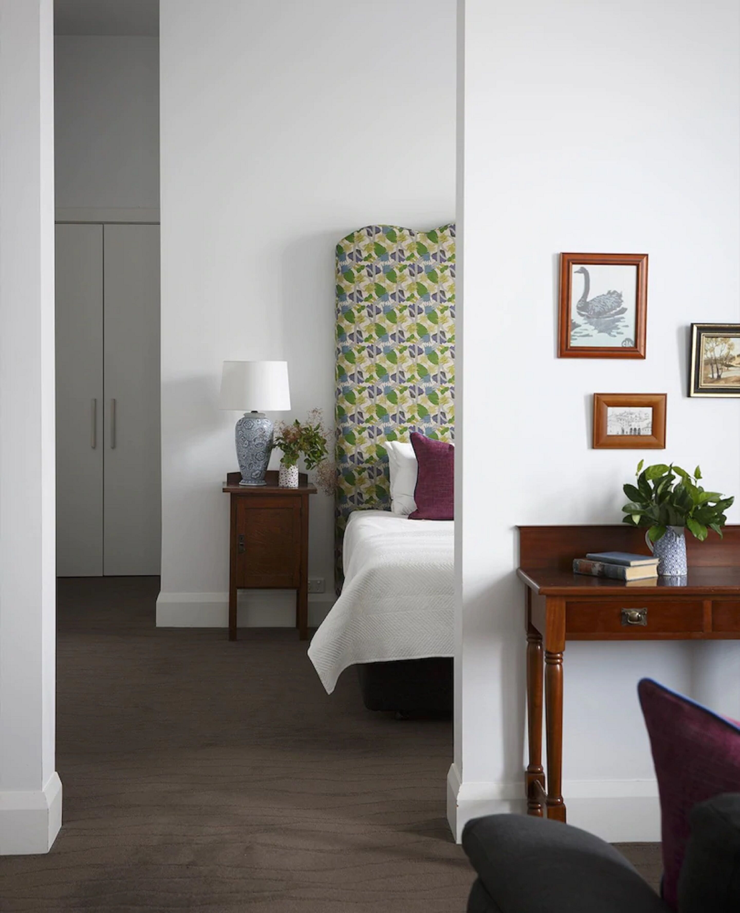 Quarto com cama, mesa de cabeceira com abajur, escrivaninha com planta e parede com quadros.