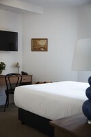 Quarto de hotel com cama, escrivaninha, cadeira, abajur e um quadro na parede.