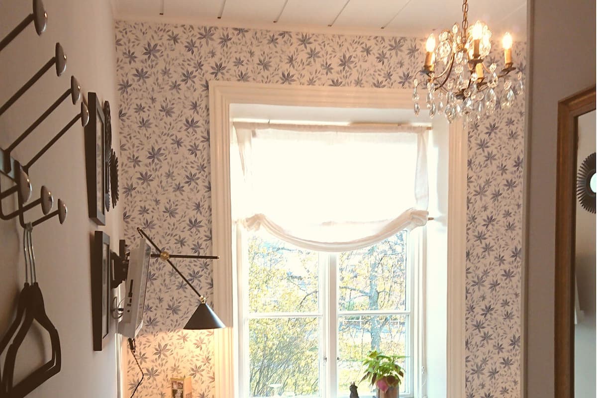 Quarto com escrivaninha, cadeira, janela com vista para árvores, lustre e parede com padrão.