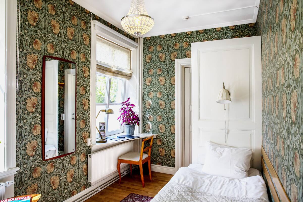 Quarto com papel de parede floral, uma cama, uma escrivaninha com cadeira, uma janela com vista e um lustre.
