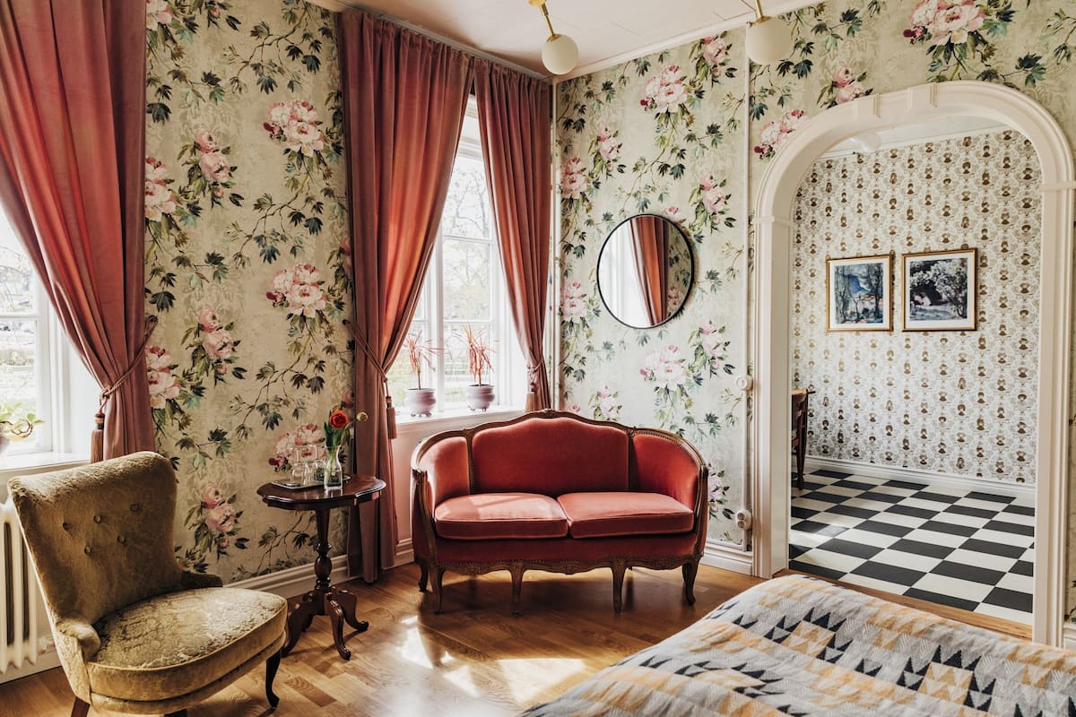 Um quarto com papel de parede floral, um sofá vermelho, uma poltrona vintage, um espelho redondo e um chão xadrez.