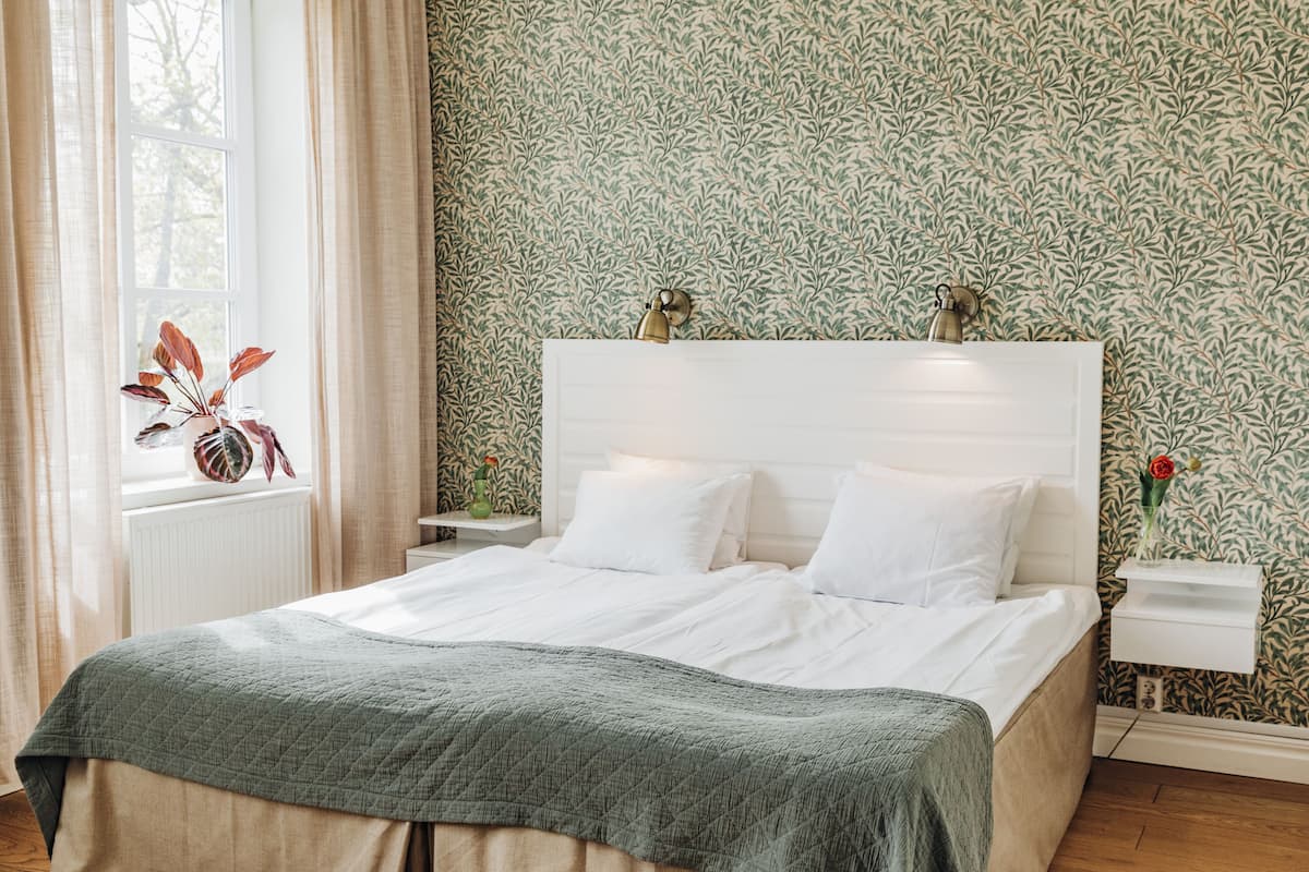 Cama bem arrumada com lençóis brancos, um edredom verde e saia bege. Há uma janela com cortinas translúcidas, um vaso com flores e uma parede com padrão de folhas.