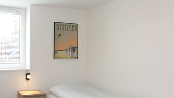Quarto pequeno e limpo, com uma cama de solteiro, mesa de cabeceira, luminária na parede e janela com cortinas.