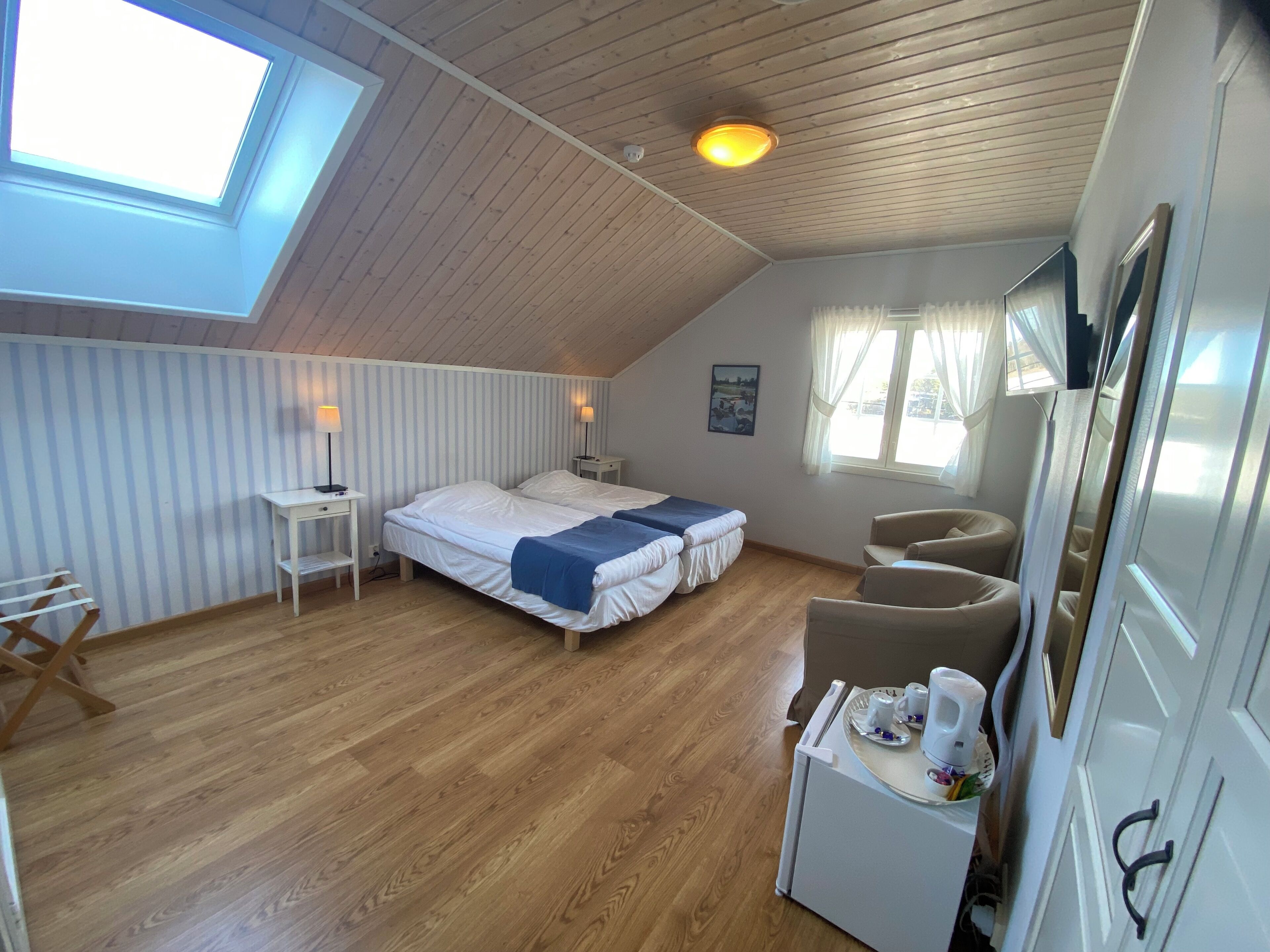 Quarto com teto inclinado, piso de madeira, cama com roupa de cama branca, escrivaninha com abajur, uma cadeira e uma pequena geladeira com itens por cima.