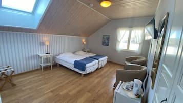 Quarto com teto inclinado, piso de madeira, cama com roupa de cama branca, escrivaninha com abajur, uma cadeira e uma pequena geladeira com itens por cima.