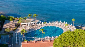 Vista aérea de um resort com uma grande piscina circular, rodeada por espreguiçadeiras e palmeiras, e uma vista desimpedida do oceano.