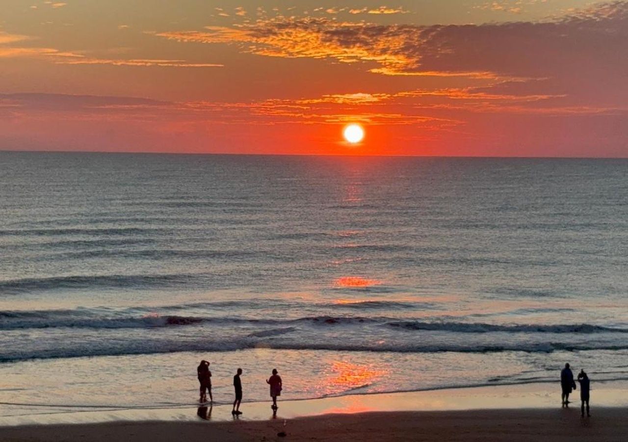 Pôr do sol sobre o oceano, com pessoas na praia.