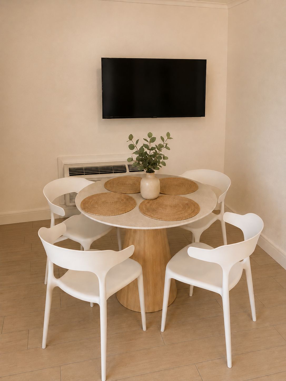Mesa redonda com base de madeira, rodeada por cadeiras brancas, uma televisão de tela plana na parede e uma pequena planta em vaso.