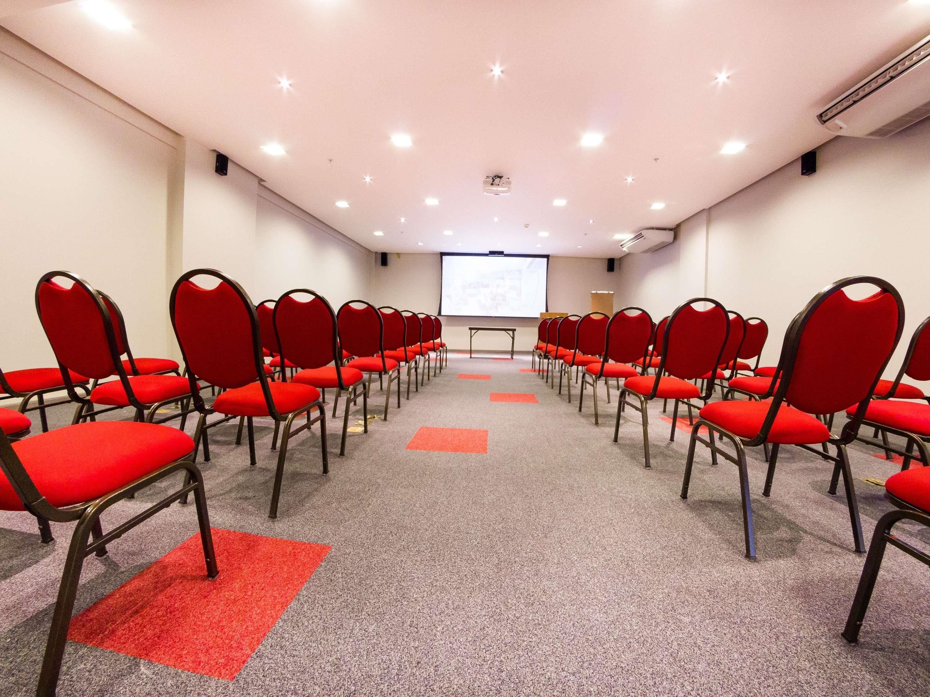 Uma sala de conferências com fileiras de cadeiras vermelhas, piso coberto de carpete e uma tela de projetor na frente.