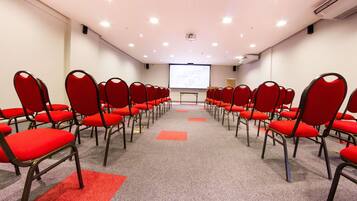 Uma sala de conferências com fileiras de cadeiras vermelhas, piso coberto de carpete e uma tela de projetor na frente.