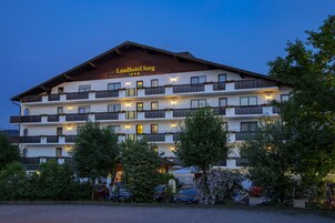Um hotel de vários andares com uma placa indicando "Landhotel Seeg". Apresenta varandas com caixas de flores, várias janelas e uma área de estacionamento na frente.