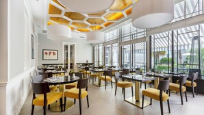 Uma sala de jantar ampla, com várias mesas e cadeiras, janelas grandes e um design de teto hexagonal único.
