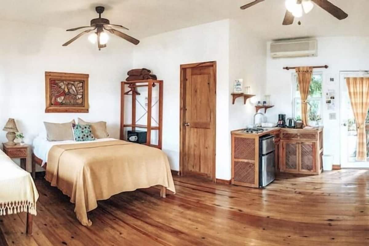 Um quarto amplo com piso de madeira, uma cama com colcha bege, um armário de madeira, um ventilador de teto e uma janela com cortinas.
